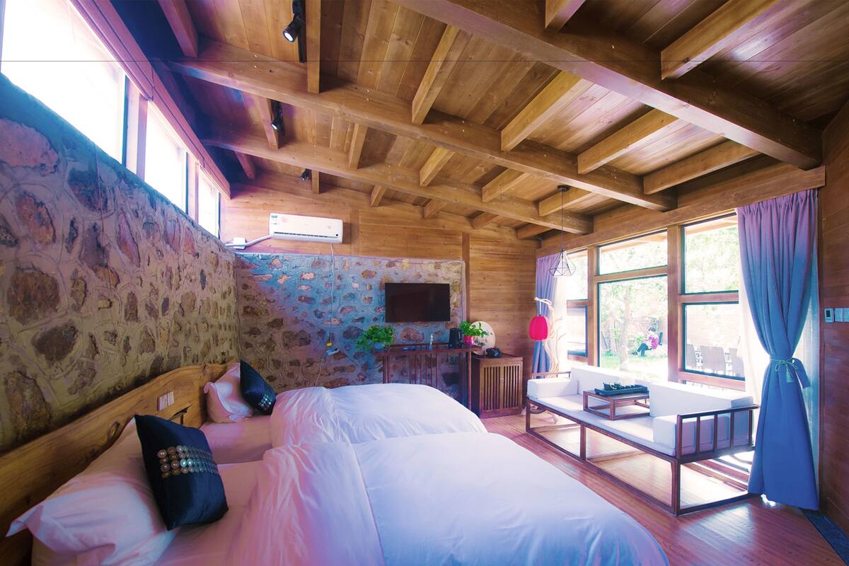 Quarto com parede de pedra, vigas de madeira no teto, cama com roupa de cama branca, uma mesinha e uma janela com cortinas.