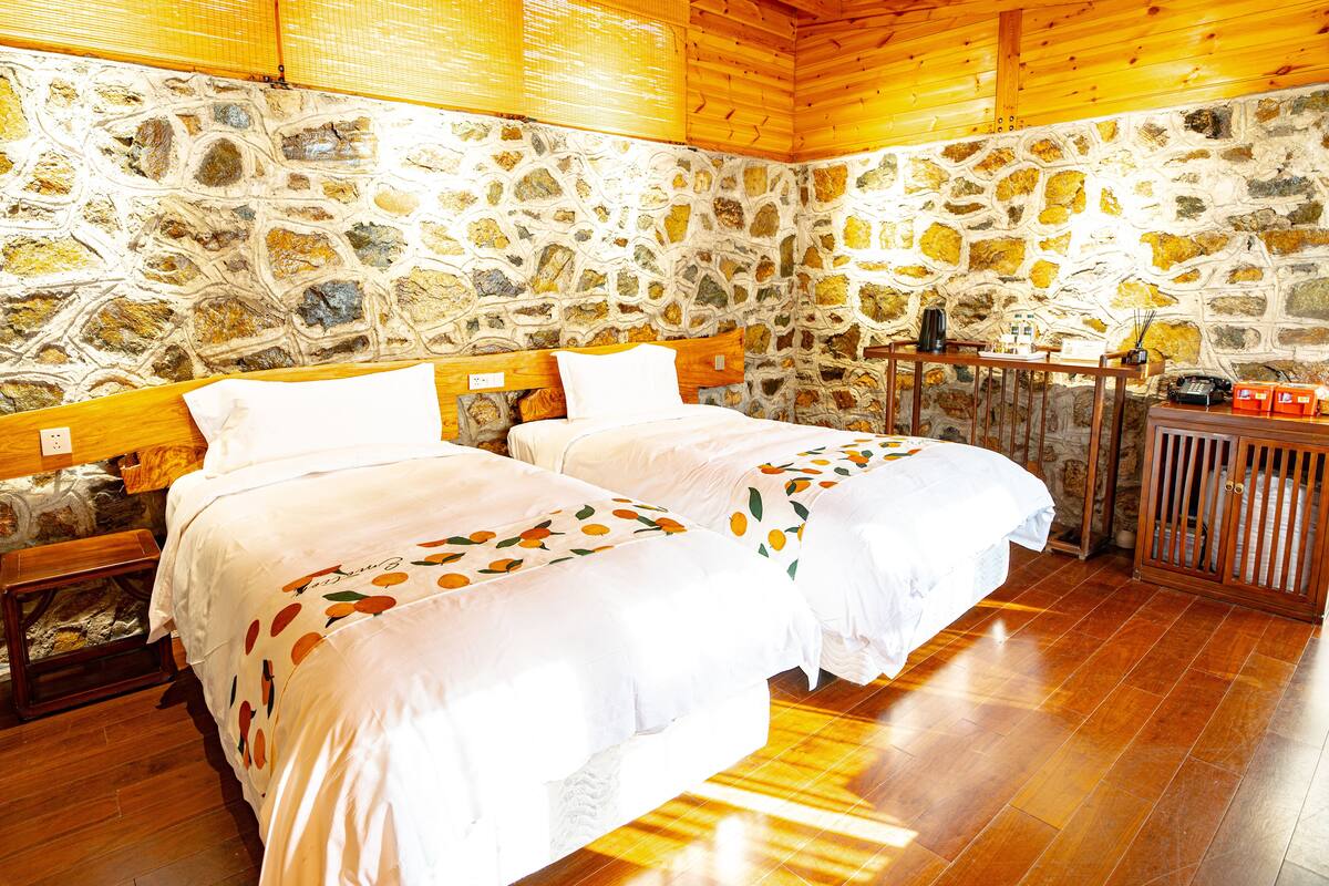 Quarto com duas camas, parede de pedra, vigas de madeira no teto e uma pequena mesa de madeira com um rádio.