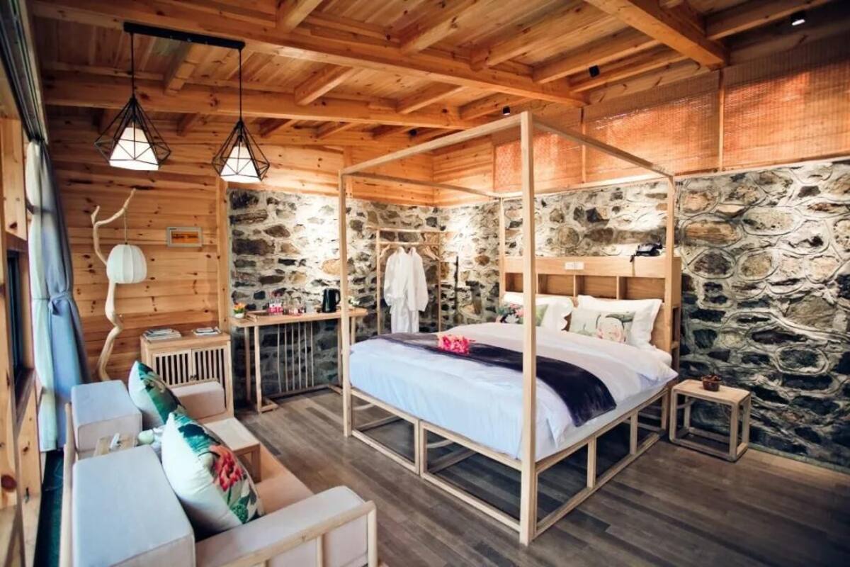 Uma cabana de madeira com parede de pedra, uma cama com dossel, uma mesa pequena e um banco.