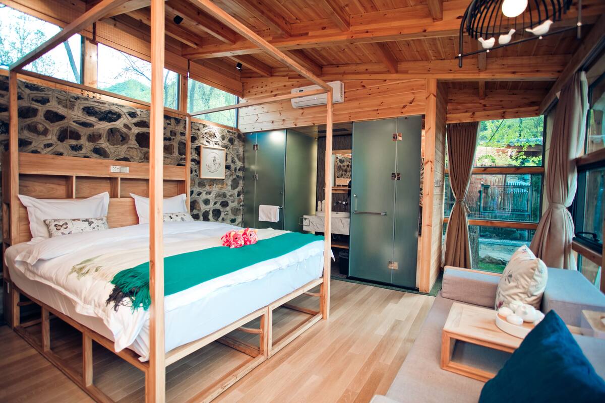 Quarto de cabana de madeira com cama, um banquinho e parede de pedra.