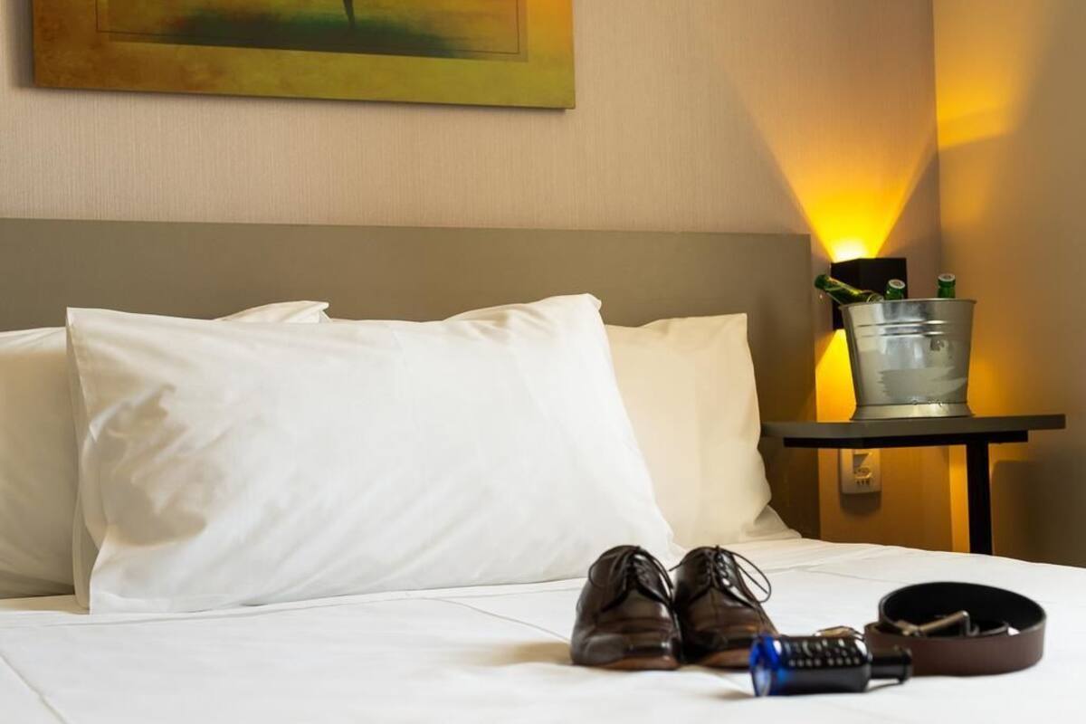 Cama bem arrumada com roupa de cama branca, um par de sapatos, um cinto e um controle remoto sobre a cama. Há uma mesa pequena ao lado da cama com um balde contendo uma planta e garrafas.