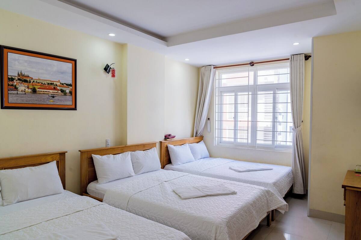 Quarto de hotel com três camas, janela com cortinas, quadro na parede e uma pequena mesa.