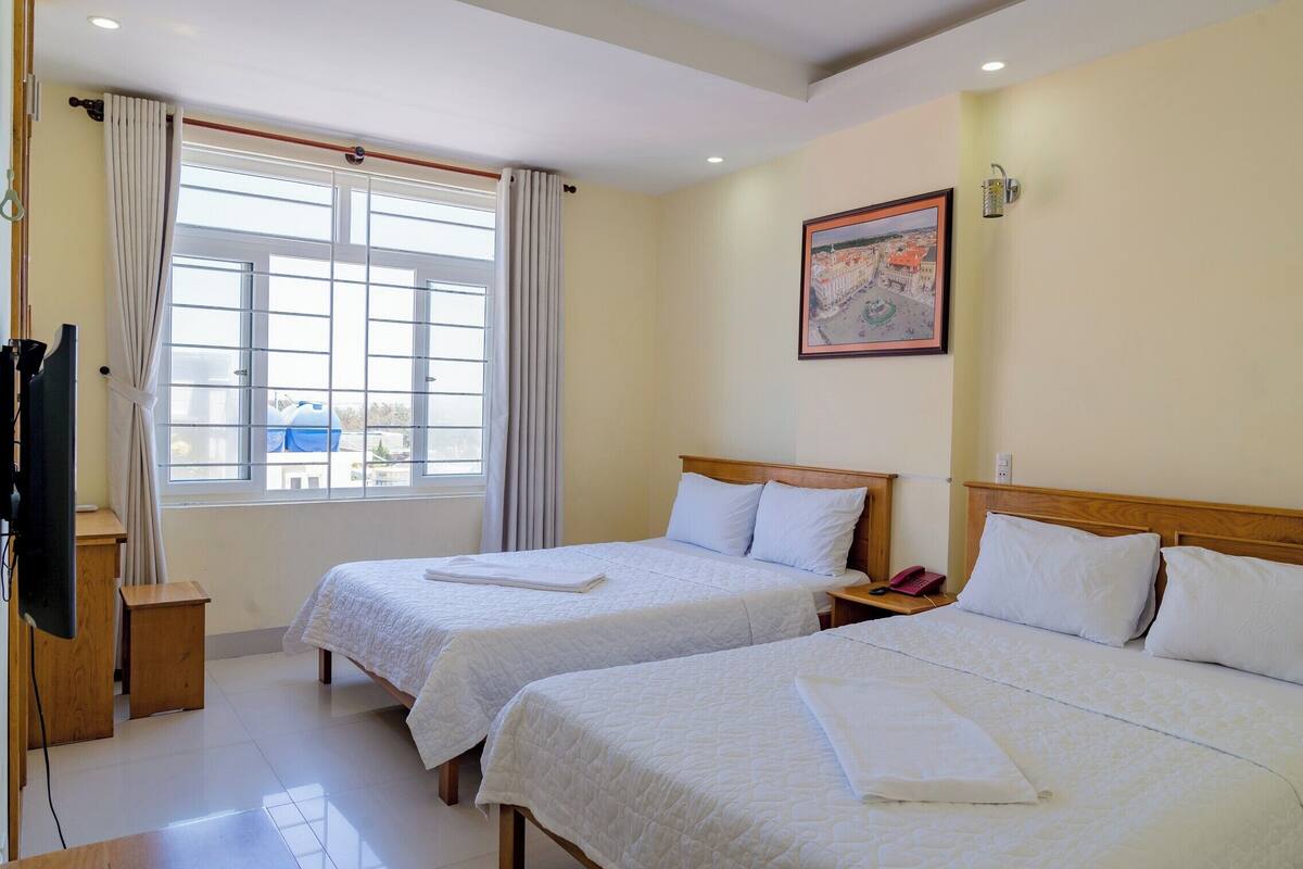 Quarto de hotel com duas camas, uma janela com cortinas, uma mesinha de cabeceira com telefone e um quadro na parede.
