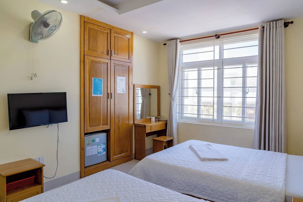 Quarto de hotel com duas camas, televisão, guarda-roupa de madeira, secretária, espelho e janela com cortinas.