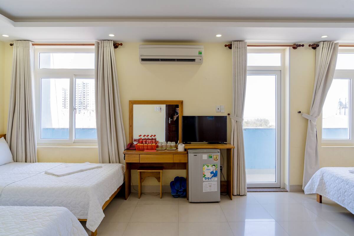Quarto de hotel com duas camas, uma escrivaninha com computador, um frigorífico e uma janela grande com cortinas.