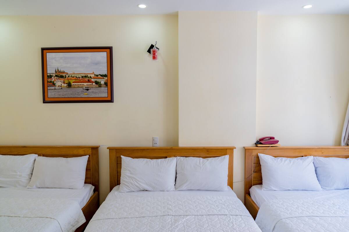 Quarto de hotel com três camas, um quadro emoldurado de uma vista urbana e um objeto vermelho pequeno na parede.