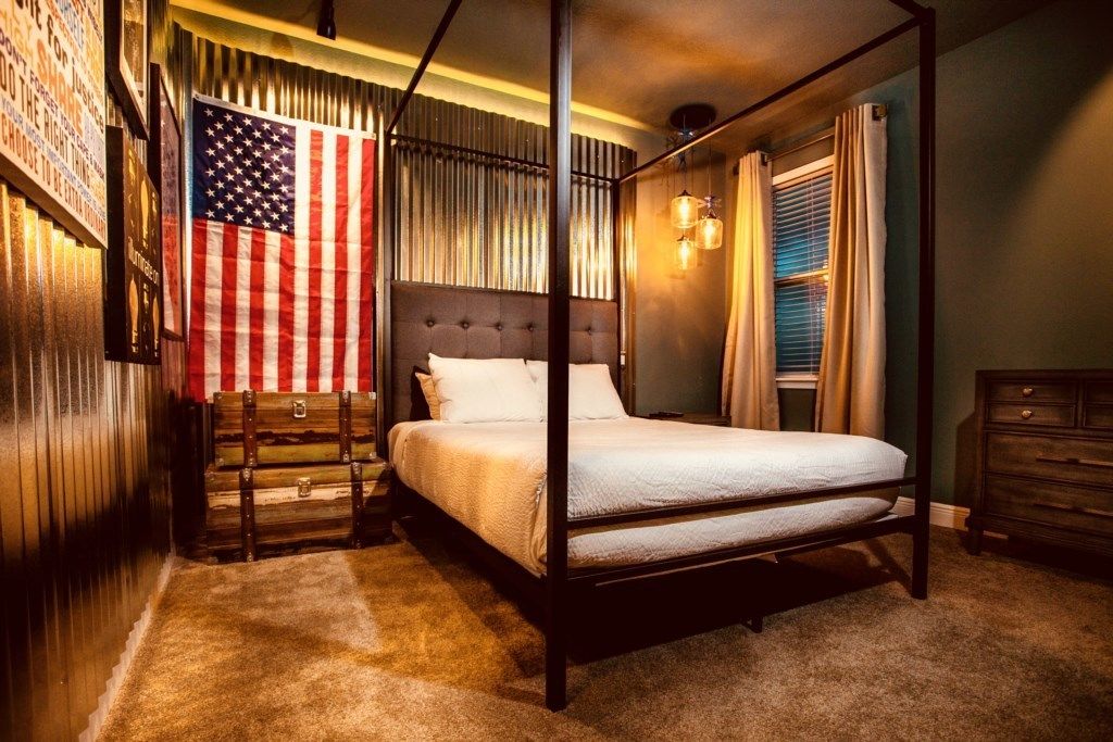 Uma cama de dossel com cabeceira estofada, uma grande bandeira americana e um conjunto de gavetas de madeira.