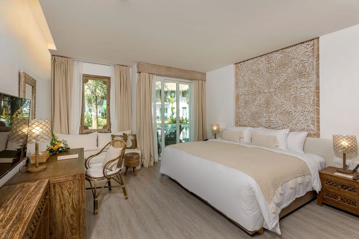 Quarto com cama grande, mesa de cabeceira de madeira, cadeira de vime, televisão e vista para a varanda.