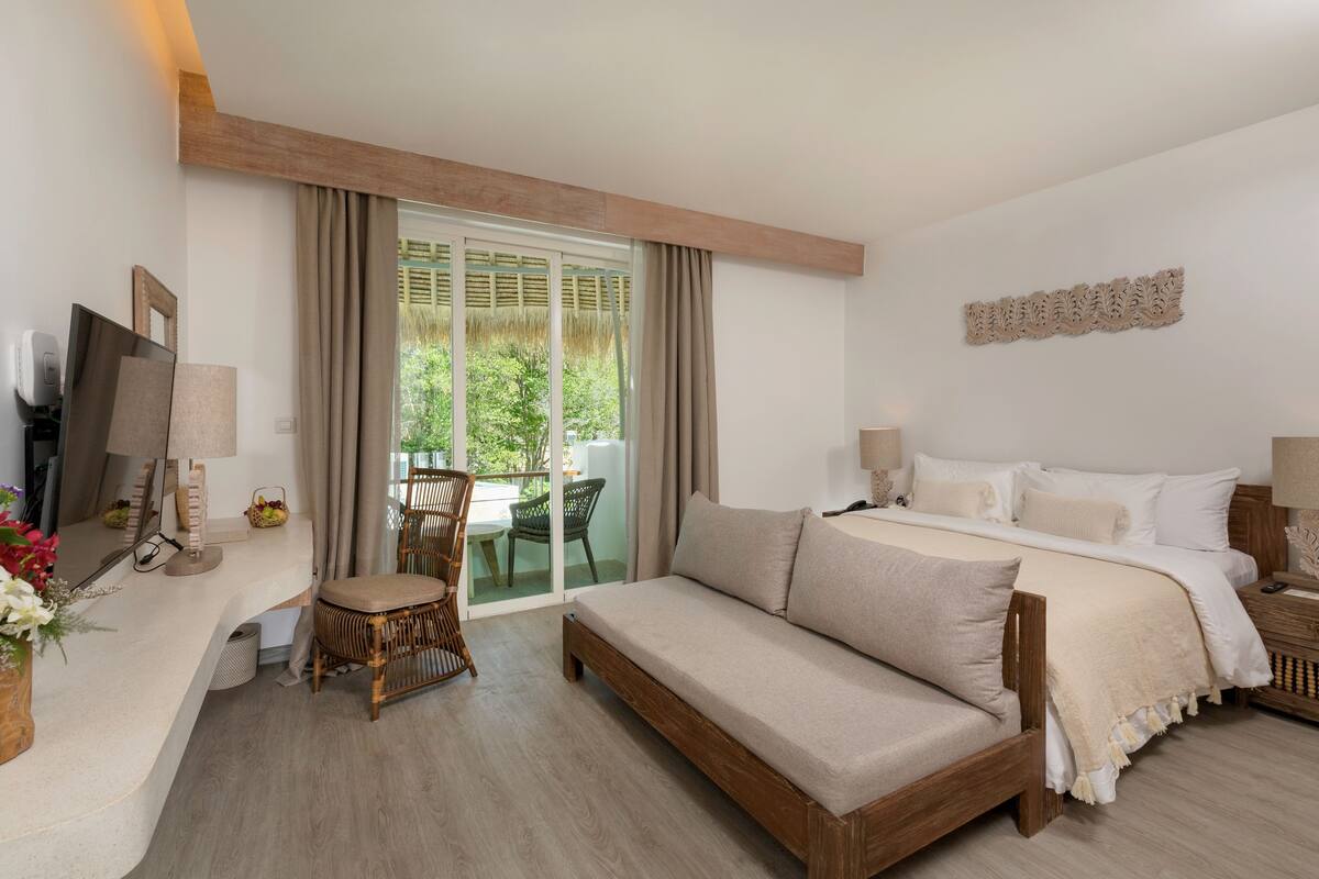 Quarto de hotel moderno com sofá-cama, televisão, área de jantar com mesa e cadeiras, e varanda com vista para o verde.