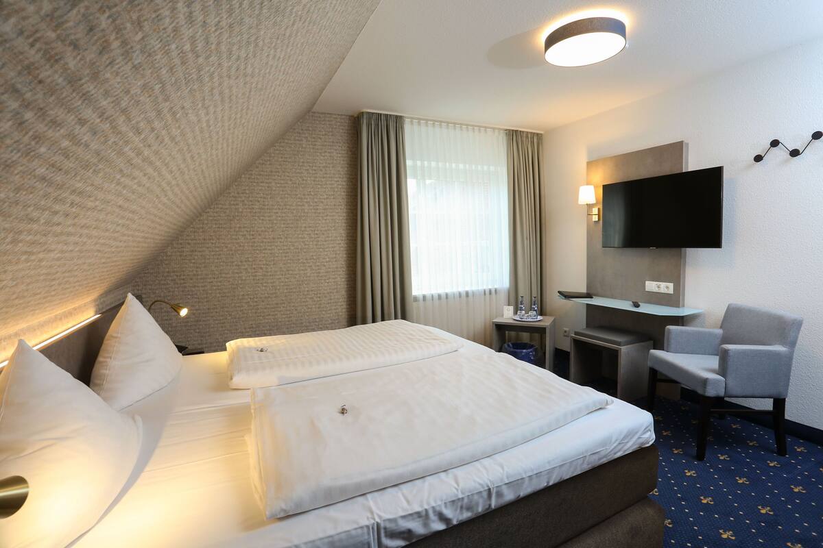 Quarto de hotel com cama, escrivaninha, cadeira, televisão e janela com cortinas.
