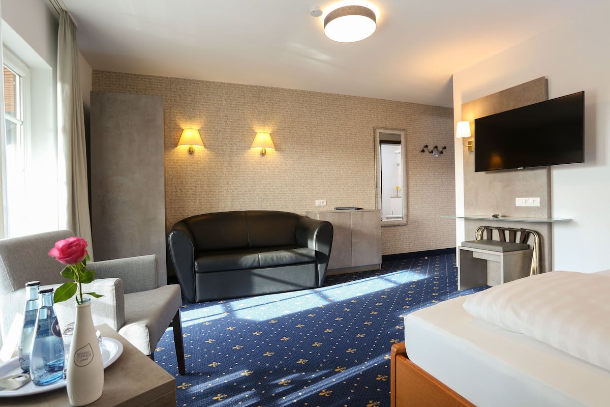 Quarto de hotel com cama, escrivaninha com vaso e garrafas de água, cadeira, televisão e TV fixada na parede.