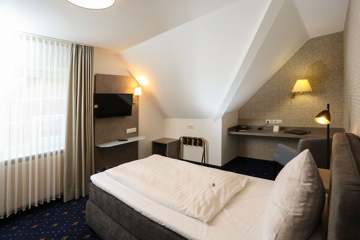 Quarto de hotel com cama, escrivaninha, televisão e janela com cortinas.
