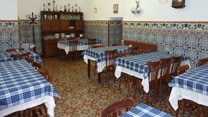 Uma área de refeições com mesas cobertas por toalhas xadrez azul e branco, cadeiras de madeira e uma parede de azulejos com uma moldura decorativa.