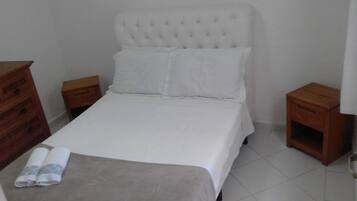 Cama bem arrumada com lençóis brancos em quarto simples com móveis de madeira e paredes brancas.