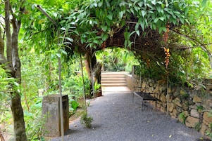 Um caminho serpenteia por um jardim, com um muro de pedra de um lado e uma estrutura de madeira do outro.