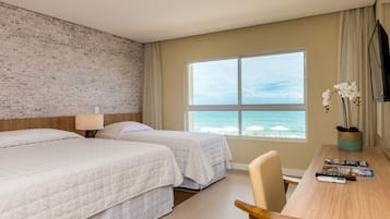 Quarto de hotel com duas camas, uma escrivaninha com cadeira, televisão e janela com vista para o oceano.