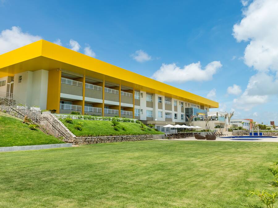 Um edifício moderno com fachada amarela, várias varandas e um pátio ajardinado com mesas e cadeiras ao ar livre.