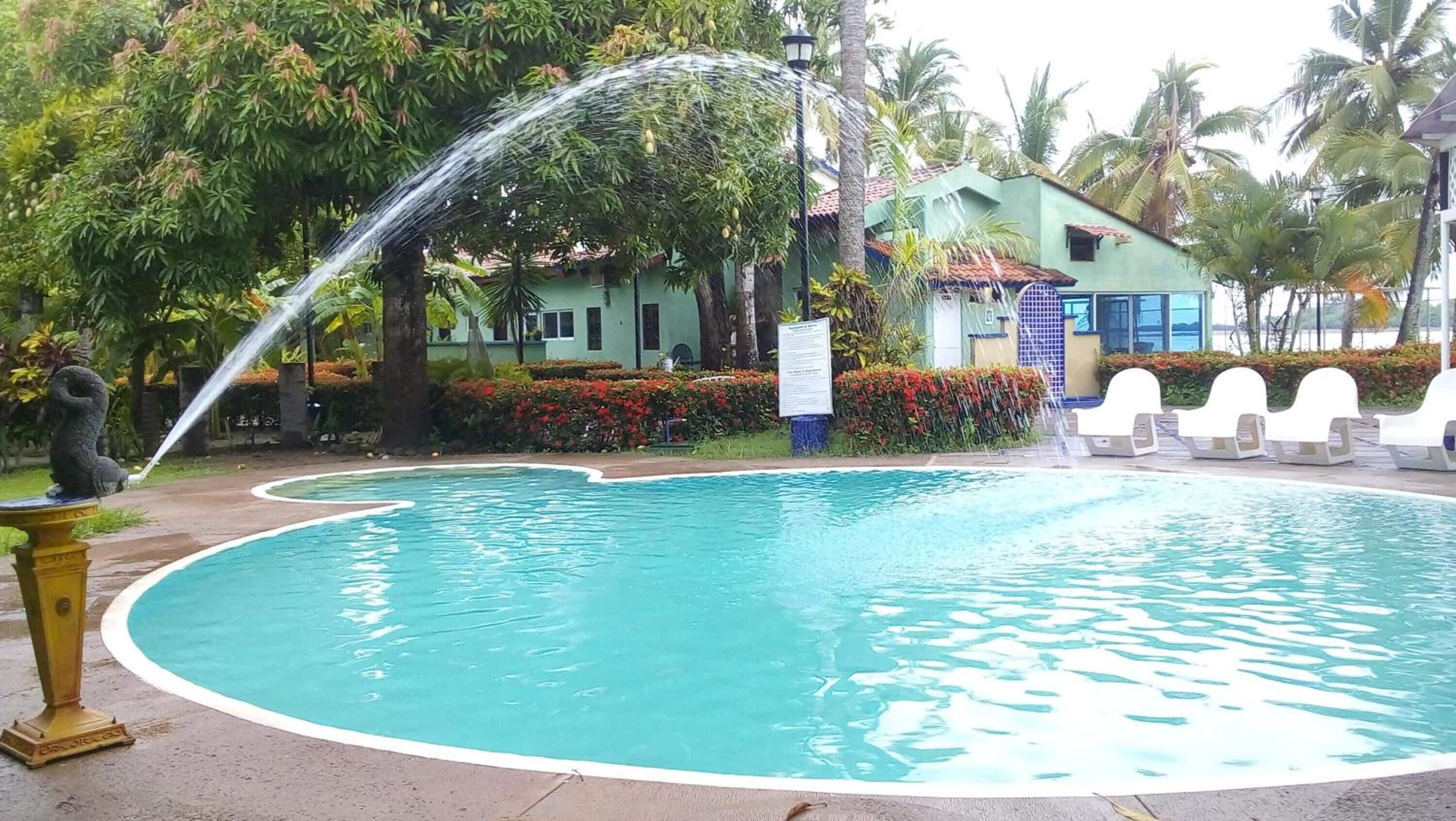 Piscina com fonte de água, cercada por vegetação exuberante e um edifício ao fundo.