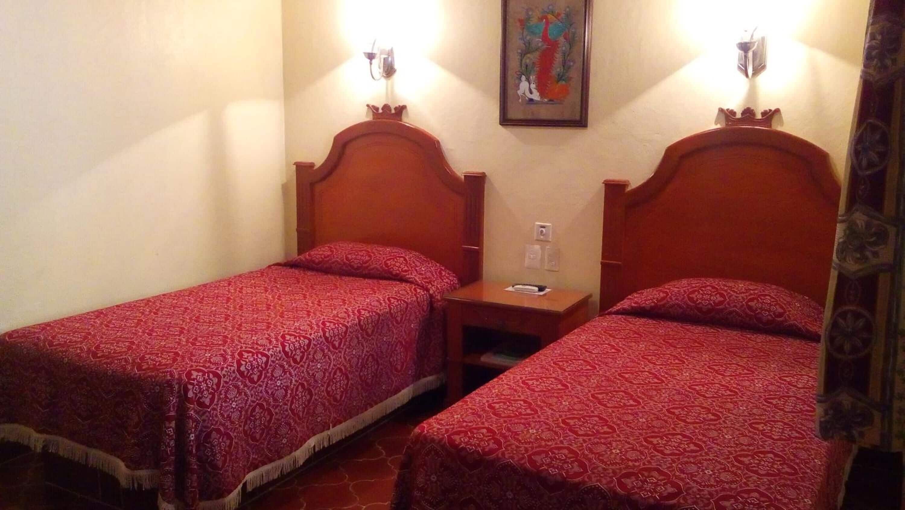 Um quarto com duas camas de madeira, cada uma com colcha e almofadas estampadas em vermelho. Há uma mesa de cabeceira entre as camas, com um abajur e uma pequena mesa.