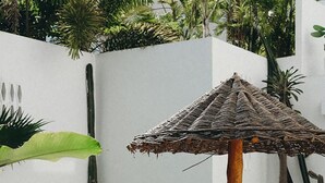 Uma área de piscina com espreguiçadeiras, um guarda-sol de palha e plantas em vasos.
