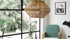 Uma sala de estar moderna com uma janela grande, uma poltrona verde, uma mesa de centro de vidro e duas luminárias pendentes de vime.