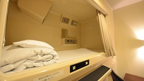 Uma cama de solteiro com travesseiro, uma mesinha de cabeceira, um pequeno prateleira e uma janela com cortinas.