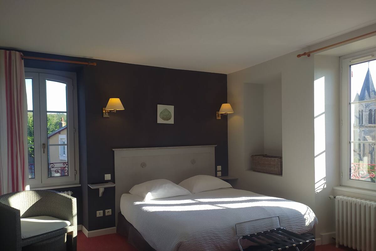 Quarto de hotel com uma cama, duas luminárias na parede, uma janela com vista para um edifício e uma cadeira.