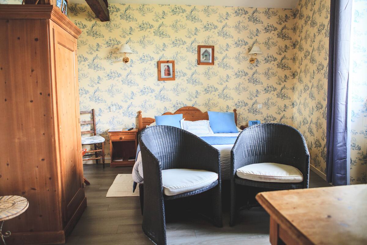 Quarto com cama, duas cadeiras de vime, mesa de madeira, guarda-roupa e mesa de cabeceira com abajur.