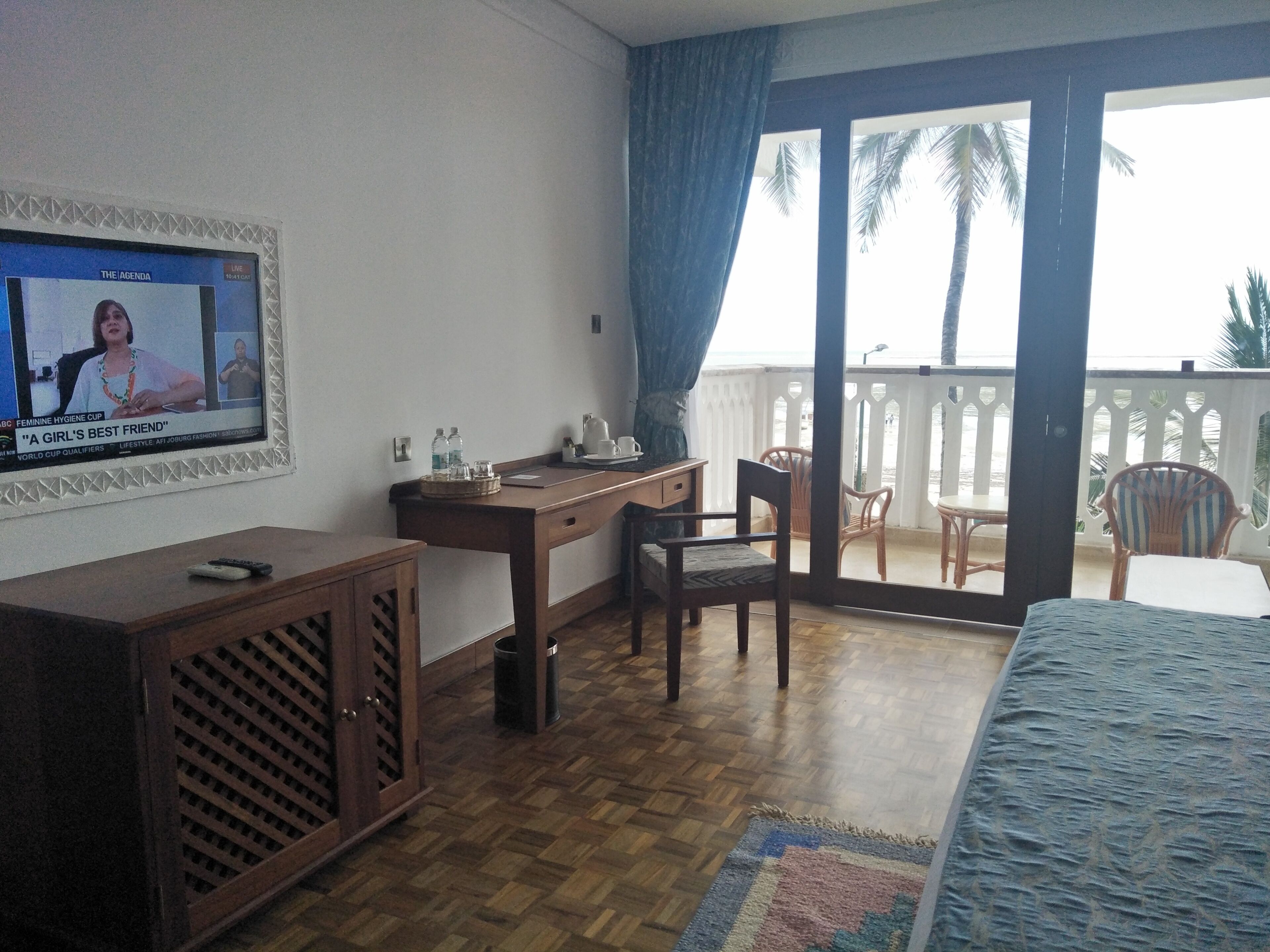 Quarto de hotel com uma escrivaninha de madeira, uma televisão exibindo um programa, uma cama e uma varanda com vista para palmeiras.