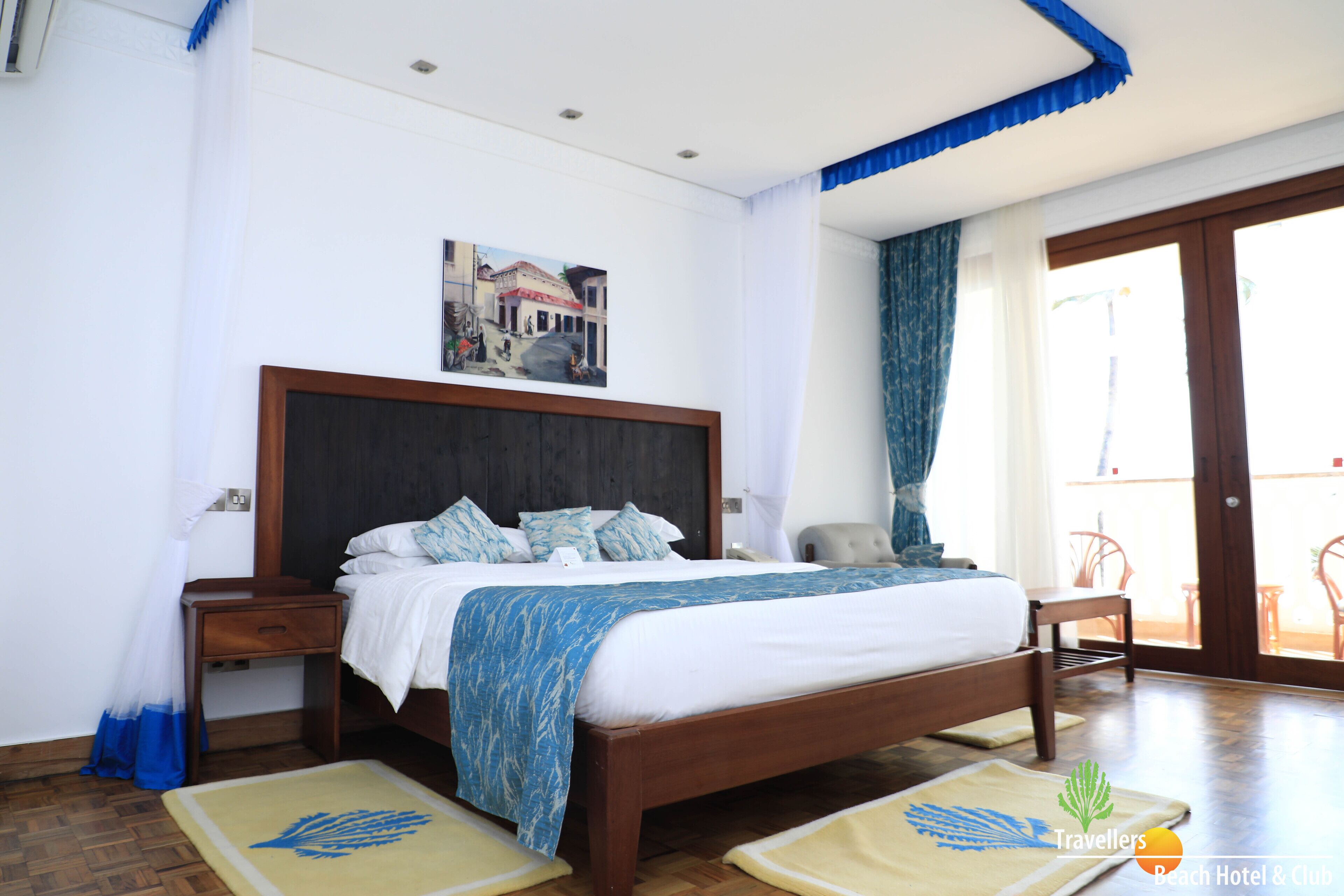 Quarto com uma cama grande, cabeceira de madeira e uma pintura de uma cena de rua. Há uma varanda com cadeiras e uma mesa na janela.