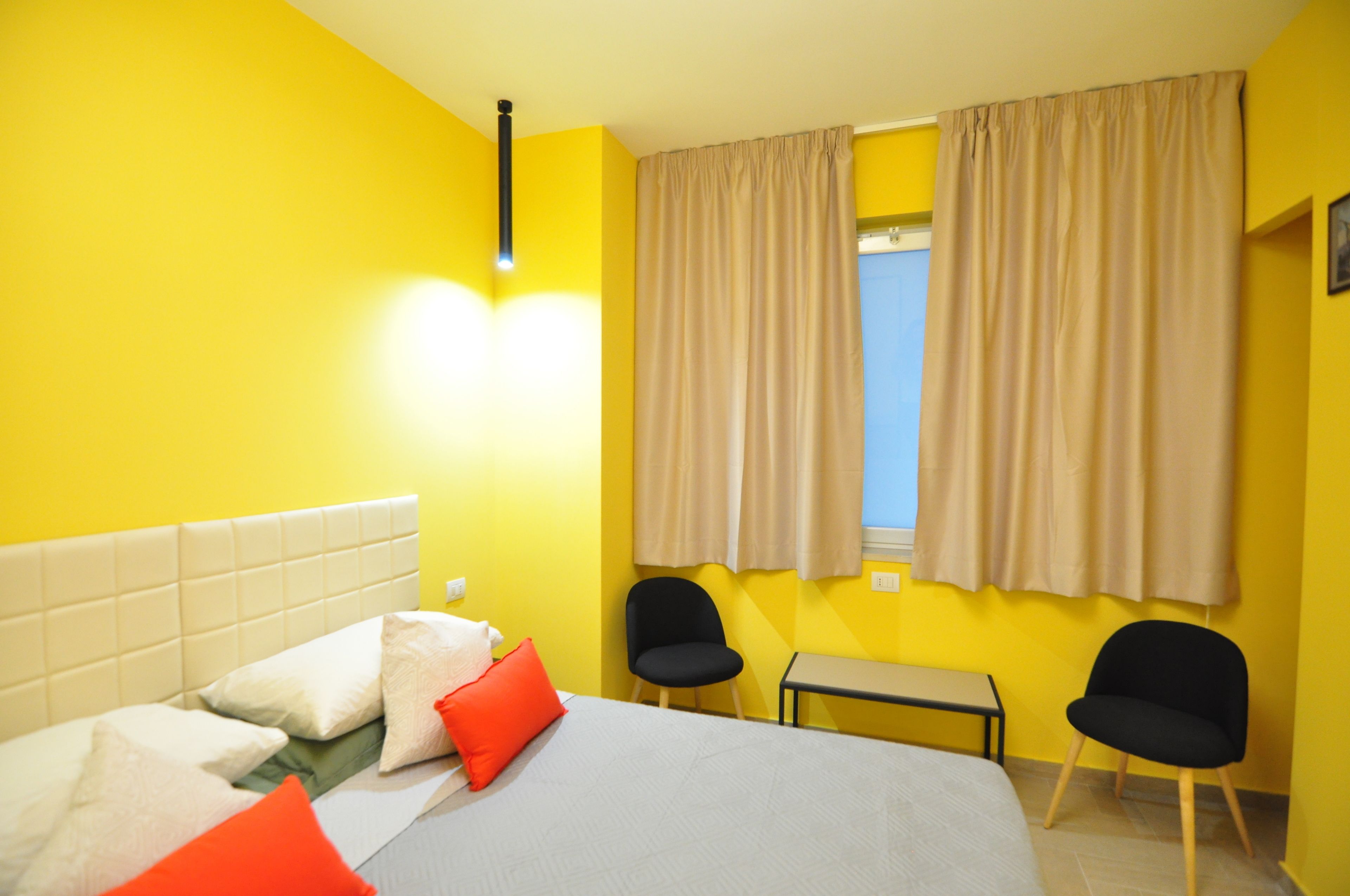 Quarto de hotel com uma cama, duas cadeiras, uma mesa pequena e uma janela com cortinas.
