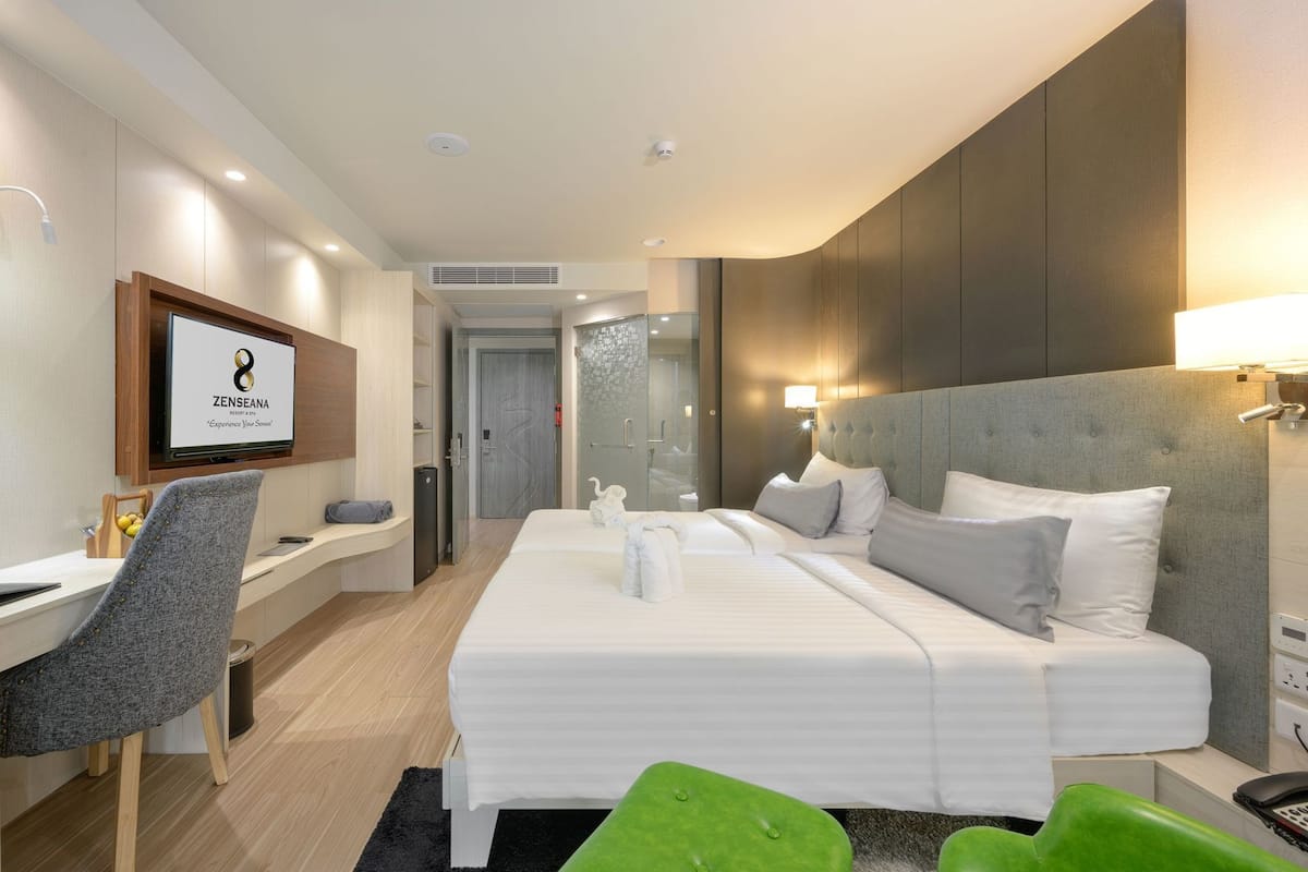 Quarto de hotel com cama, escrivaninha, cadeira, televisão e banheiro visível através de uma porta de vidro.