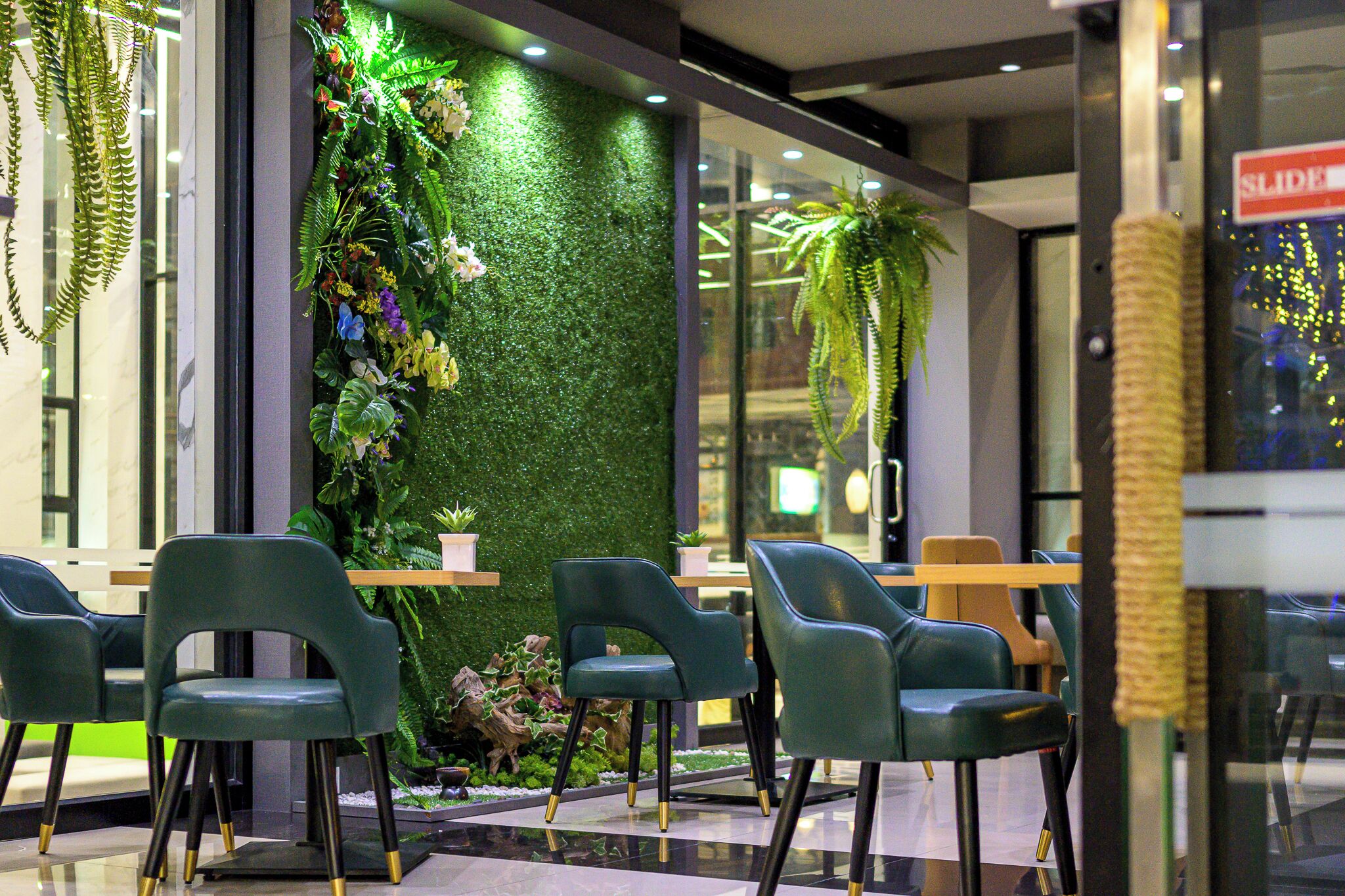 Uma área de estar moderna e interna, com cadeiras e mesas verdes, uma parede verde com plantas e uma divisória de vidro.