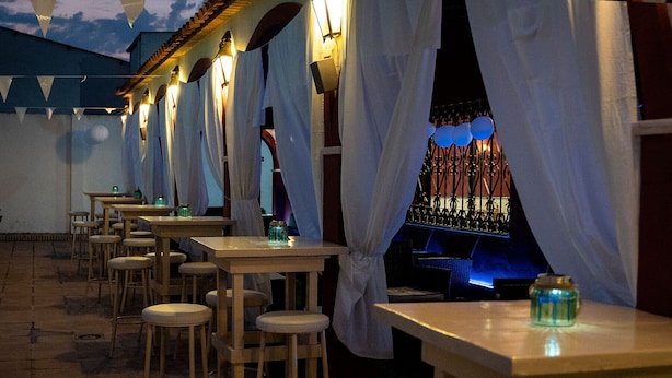 Restaurante ao ar livre com mesas e cadeiras de madeira, cortinas brancas e vista para um edifício com entrada iluminada em azul.