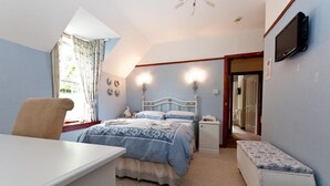 Quarto com cama, mesas de cabeceira, escrivaninha, cadeira, banco, ventilador de teto e televisão fixada na parede.
