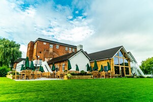 Um hotel com fachada de tijolos, um edifício branco com grandes janelas, área de estar ao ar livre com guarda-sóis verdes e um gramado.