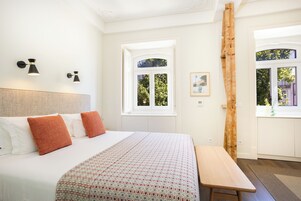 Um quarto com cama, duas luzes fixadas na parede, janela, um banco de madeira e uma porta.