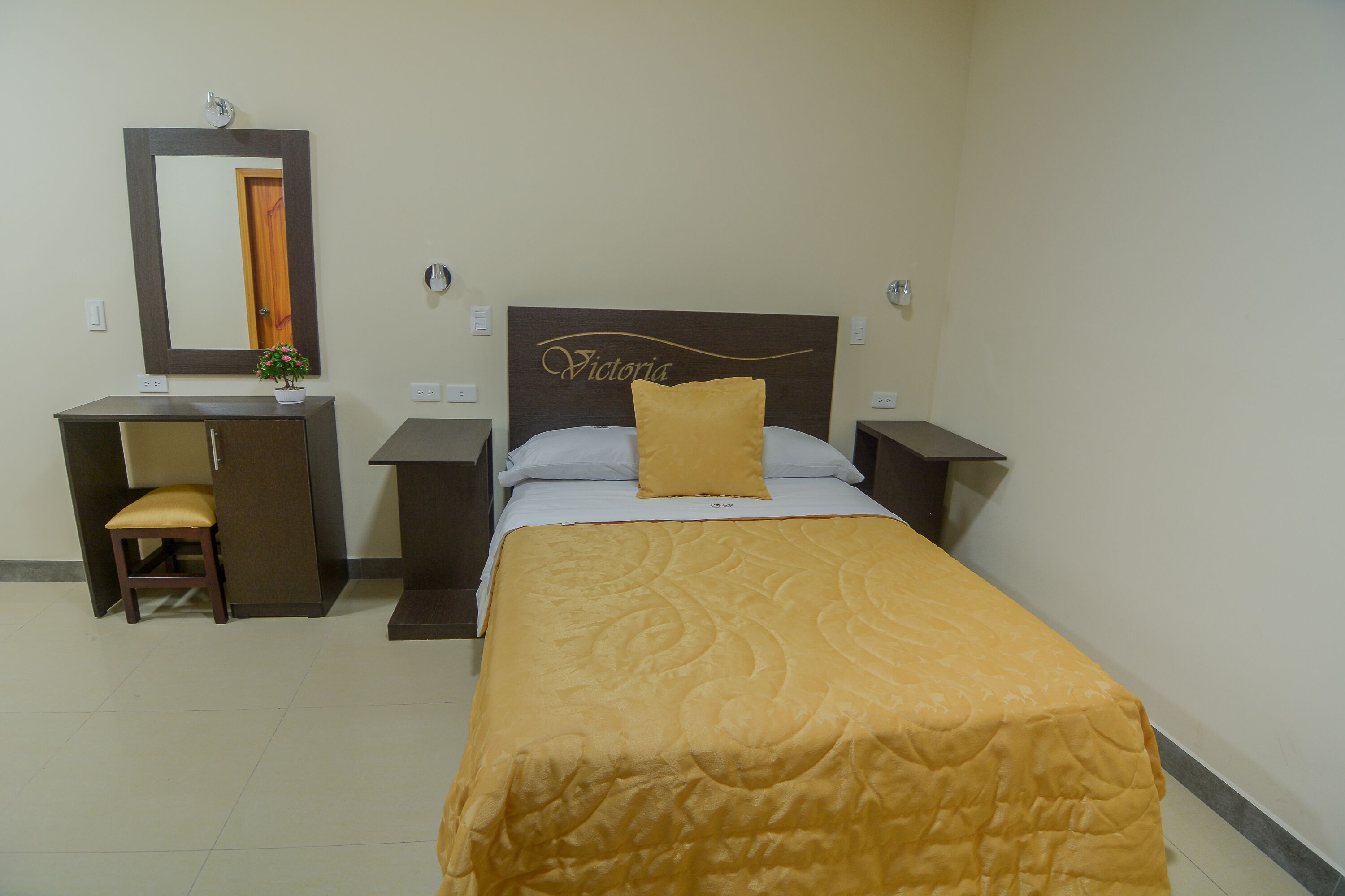 Quarto de hotel com cama, mesa de cabeceira, escrivaninha e cadeira, além de um espelho.