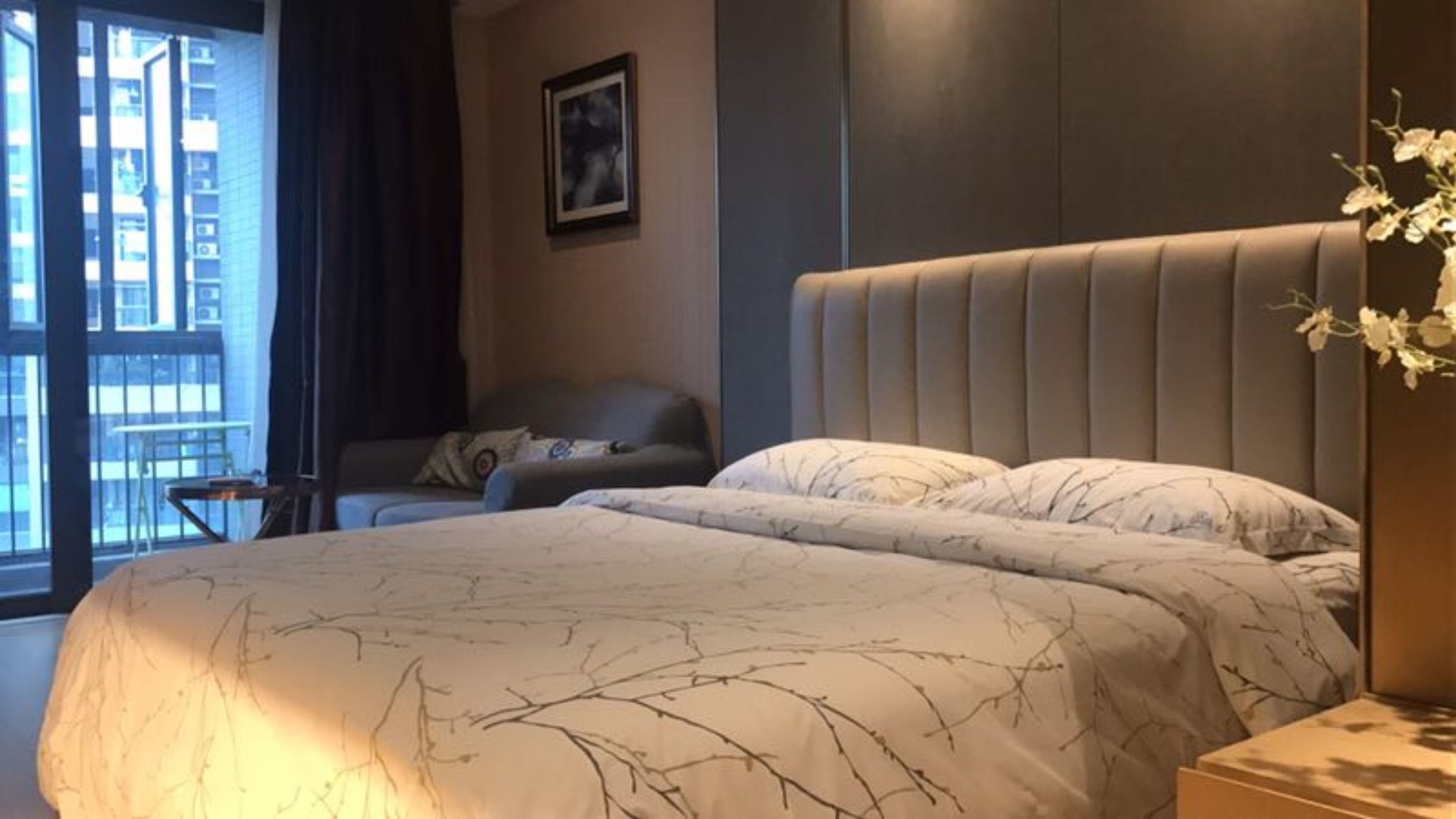 Quarto moderno com uma cama grande, mesa de cabeceira, uma cadeira e uma janela com vista para edifícios.