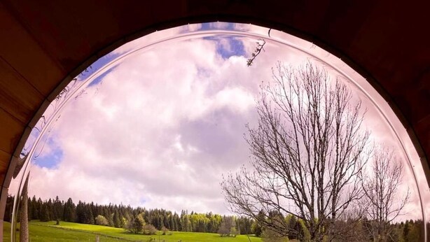 Janela circular com vista para um campo verde e árvores, bancos de madeira e um balde de madeira com uma colher.
