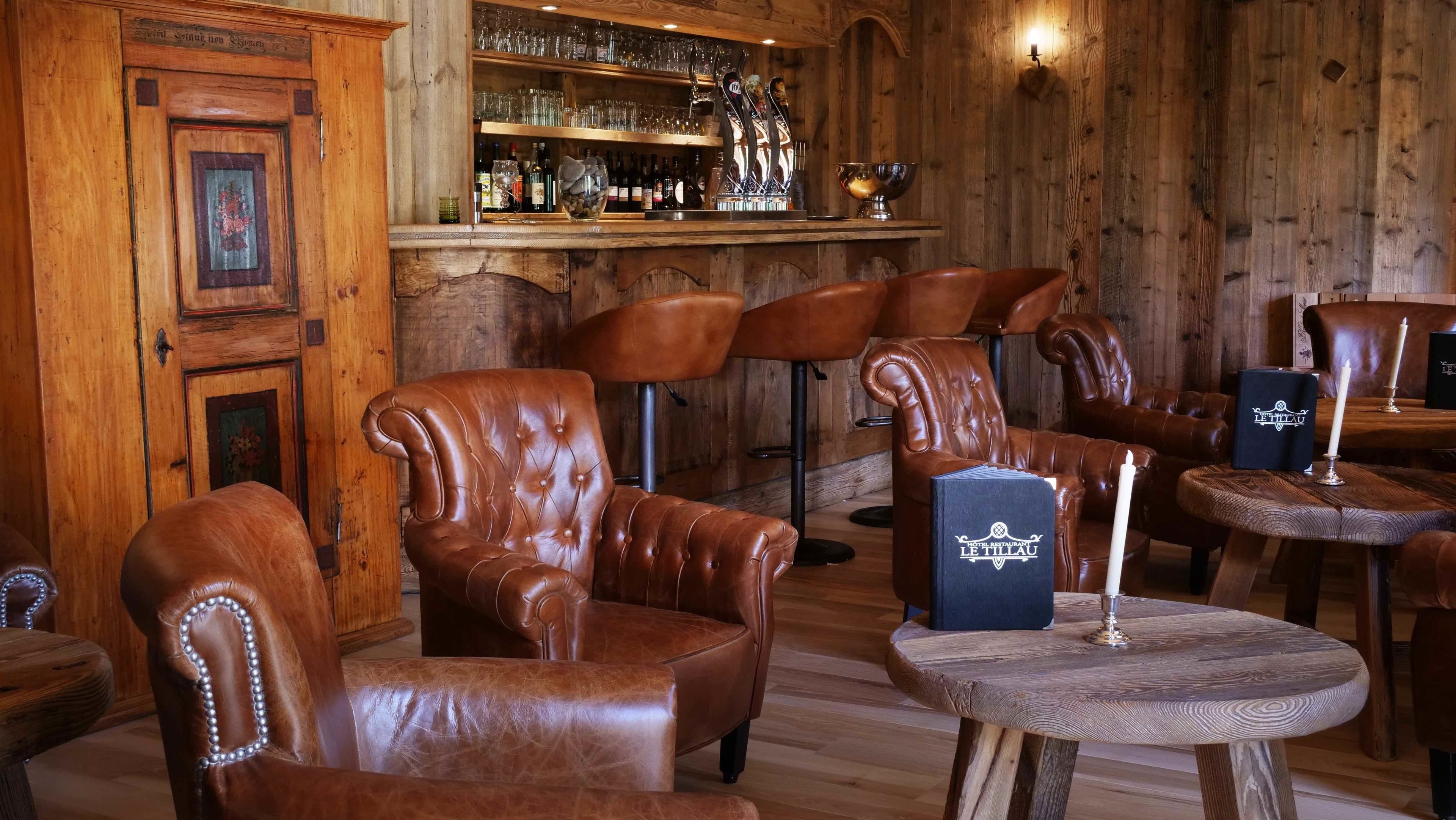 Interior em madeira com poltronas de couro, um espaço de bar com copos e garrafas, e uma mesa com uma vela.