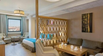 Quarto de hotel com cama, sofá, mesa de jantar com jogo de chá, divisória decorativa e vista para a cidade pela janela.