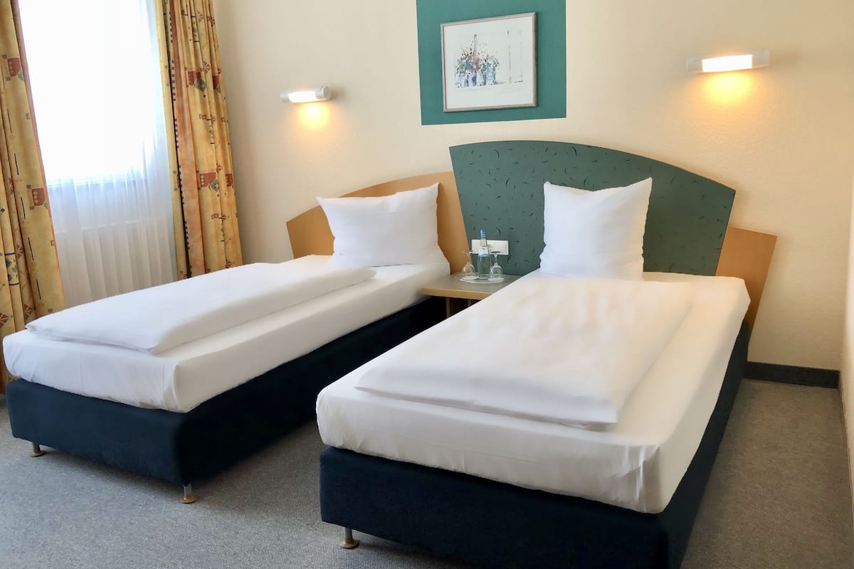 Quarto de hotel com duas camas de solteiro, uma mesa pequena com uma garrafa e copos, e um quadro na parede.