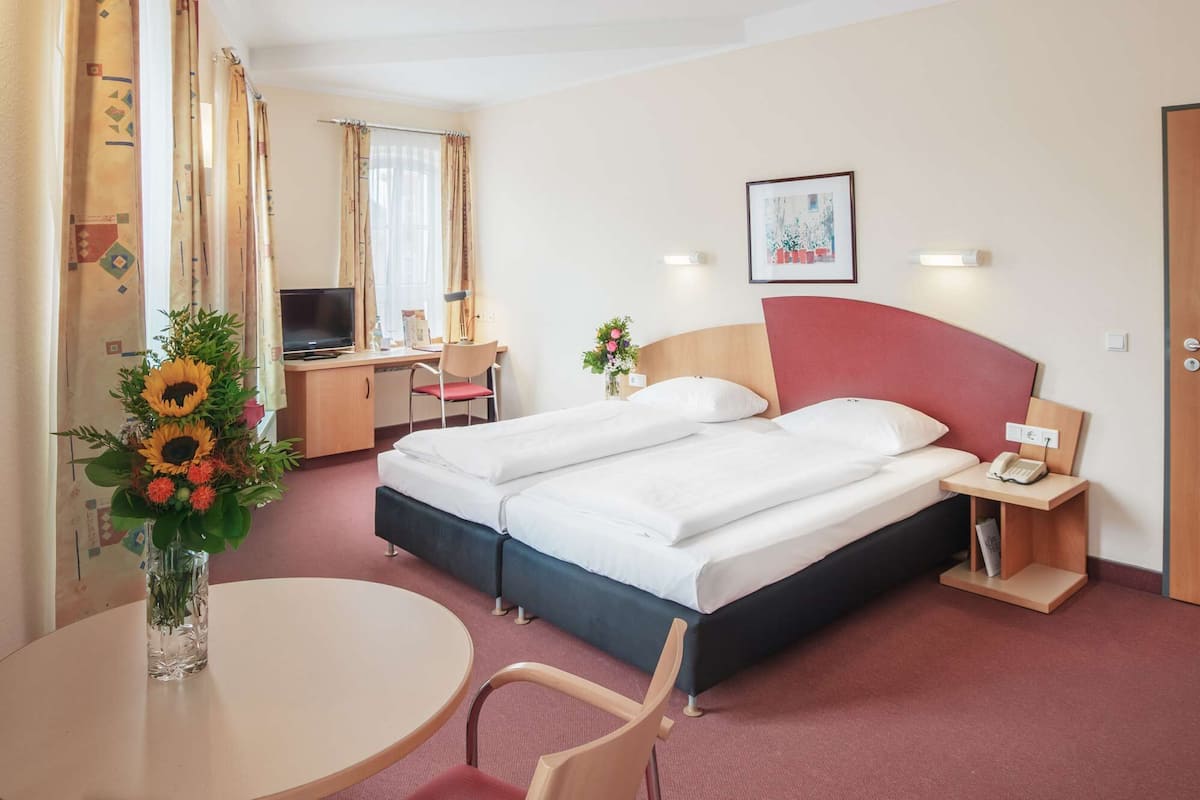 Quarto de hotel com uma cama, uma escrivaninha com um computador, uma cadeira, uma mesa com um vaso de flores e um quadro na parede.