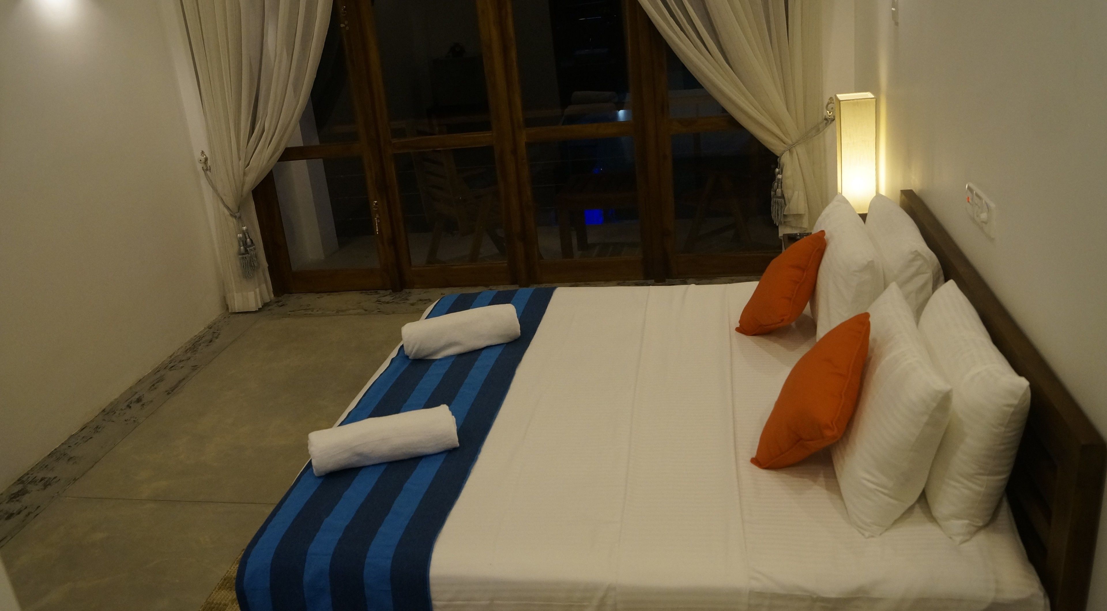 Quarto de hotel com uma cama, duas almofadas, um cobertor listrado e uma mesa de cabeceira com abajur.