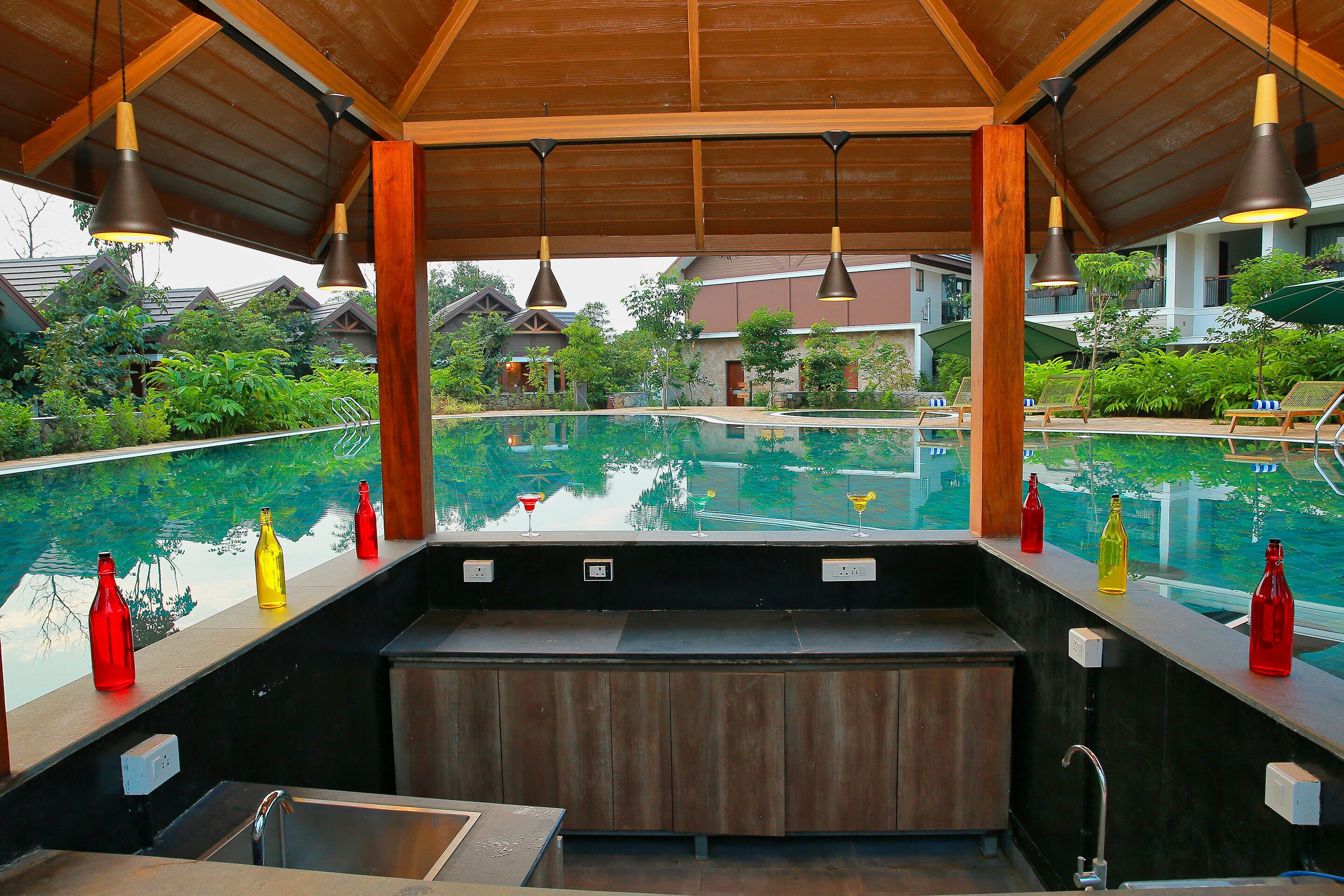 Um bar à beira da piscina com teto de madeira, vista para a piscina e garrafas coloridas no balcão.
