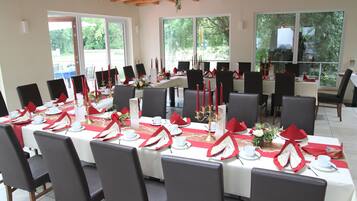 Uma sala de jantar com mesas preparadas para um evento formal, com guardanapos vermelhos, toalhas brancas e castiçais.