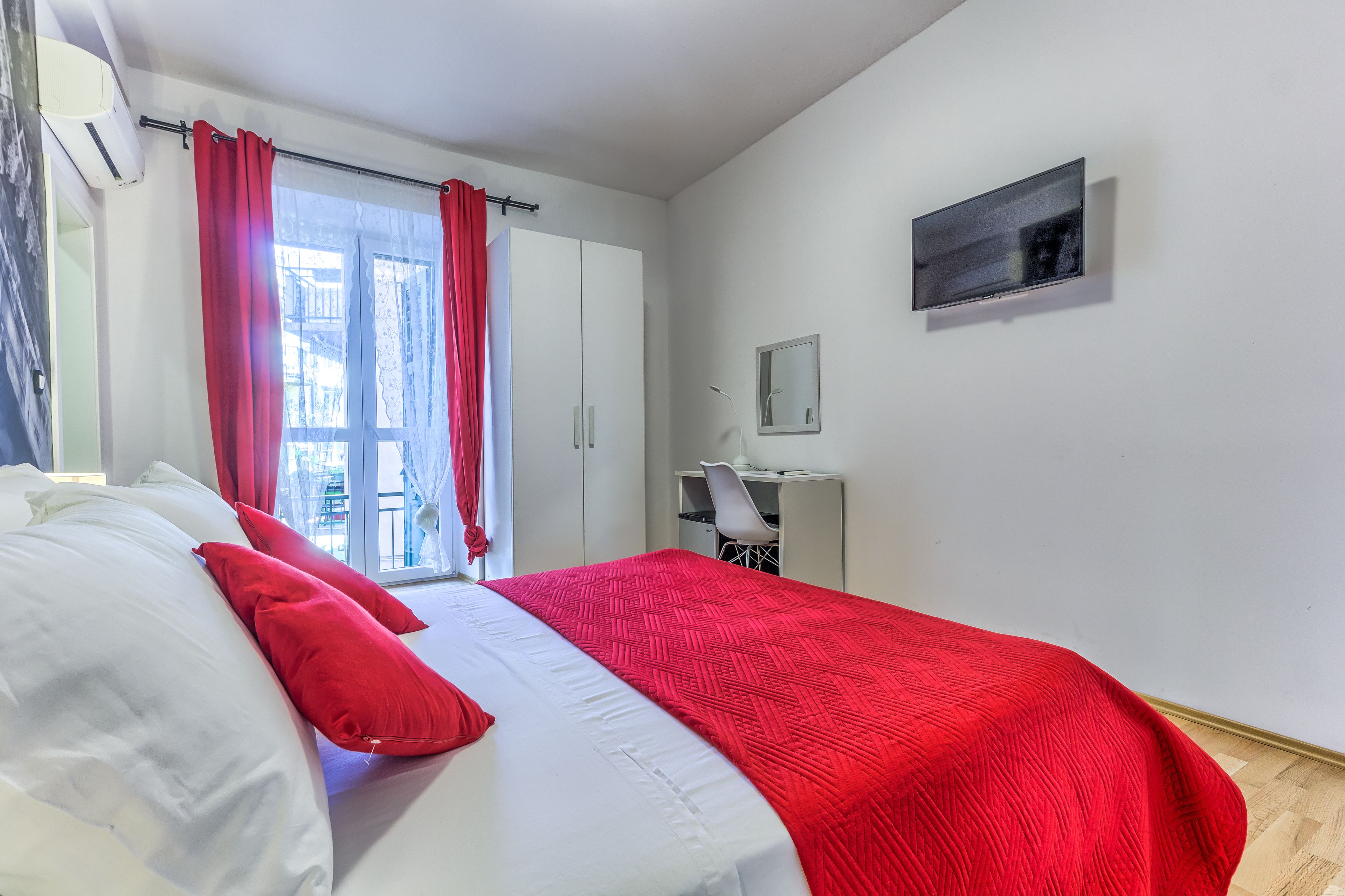 Quarto com cama, escrivaninha, cadeira, televisão e janela com cortinas.