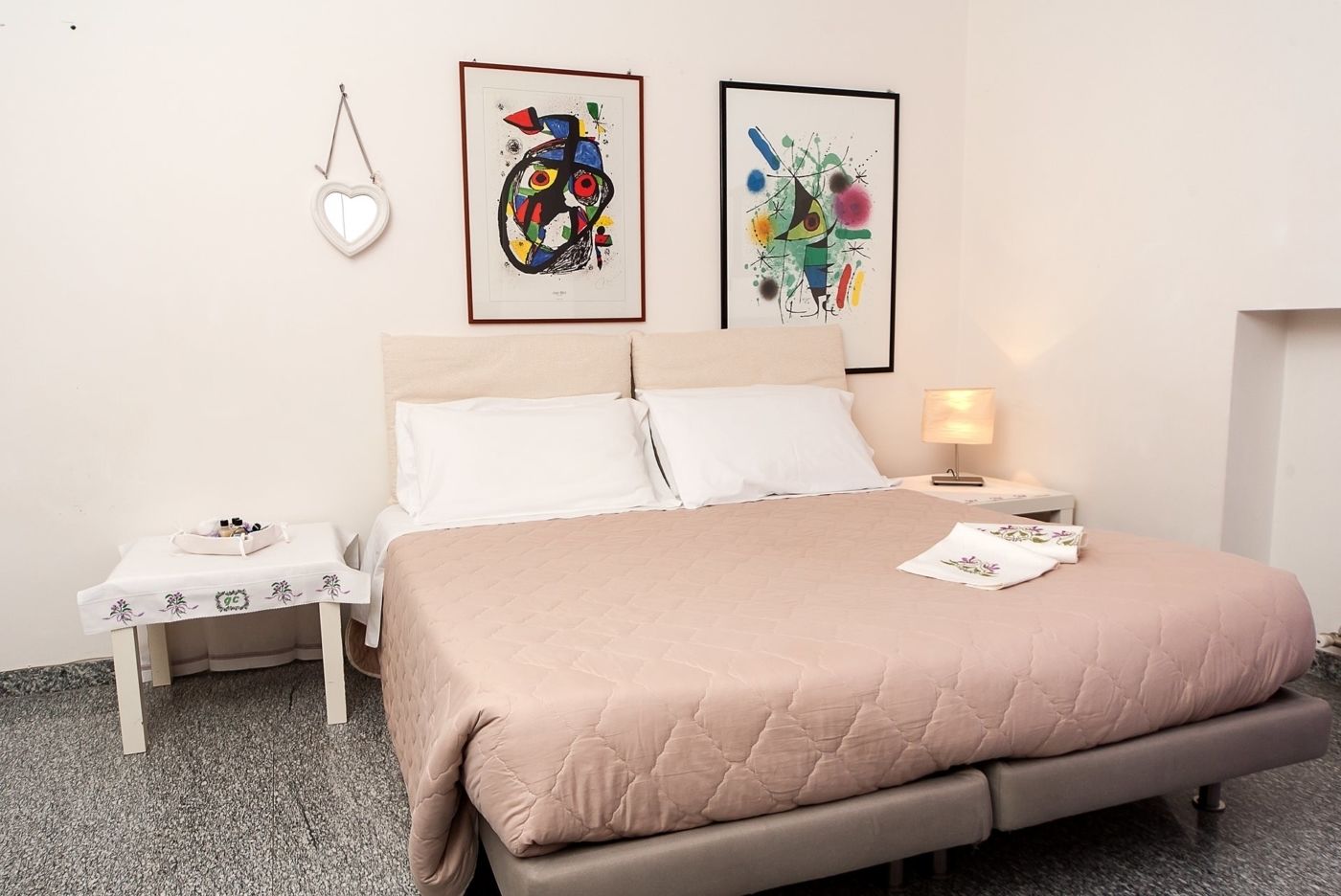 Quarto de hotel com uma cama, uma mesinha de cabeceira, uma luminária em formato de coração fixada na parede e duas pinturas abstratas emolduradas.