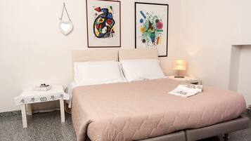 Quarto de hotel com uma cama, uma mesinha de cabeceira, uma luminária em formato de coração fixada na parede e duas pinturas abstratas emolduradas.